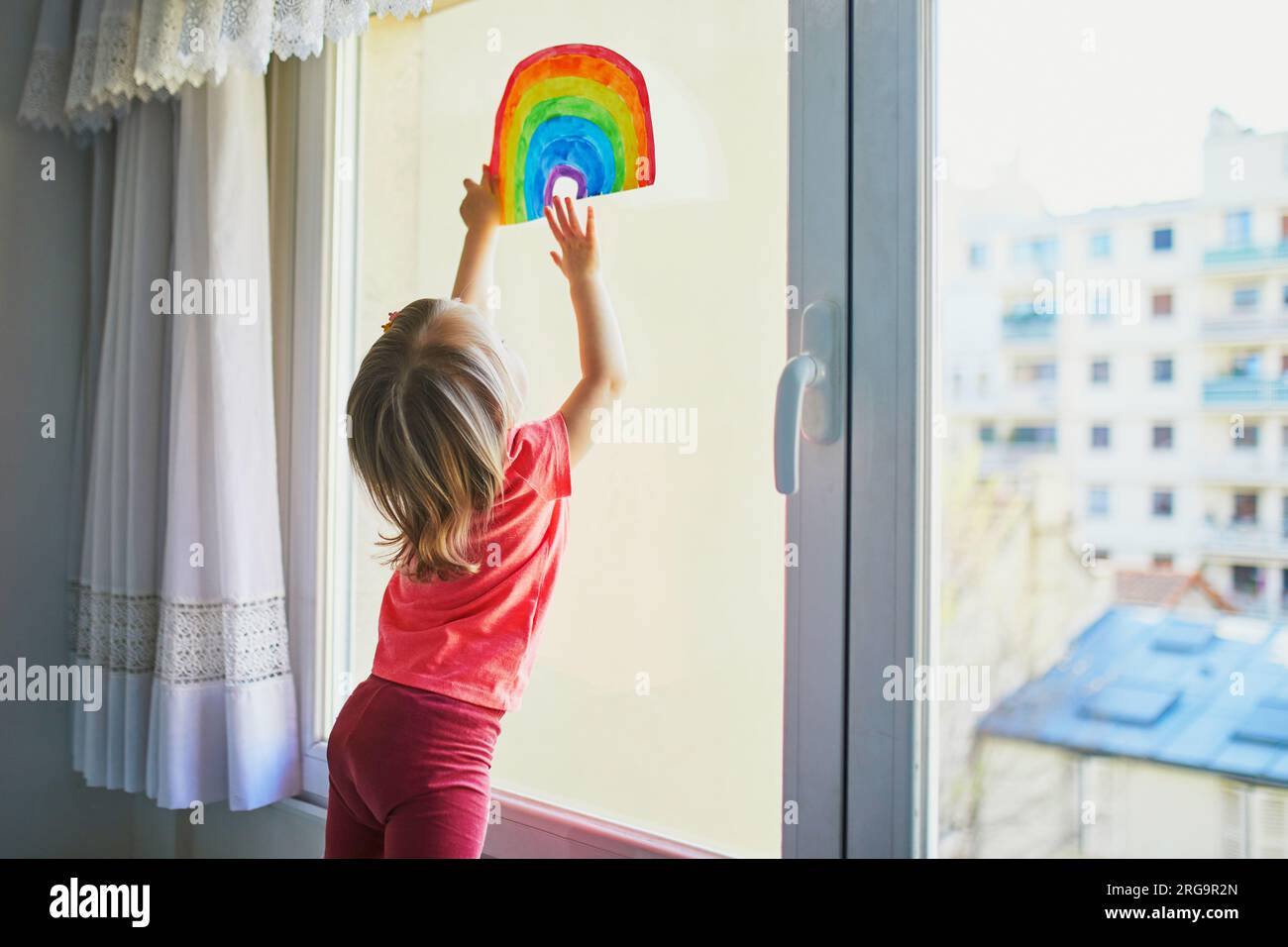 Adorable toddler girl attaching rainbow drawing to window glass as sign of hope. Creative games ...