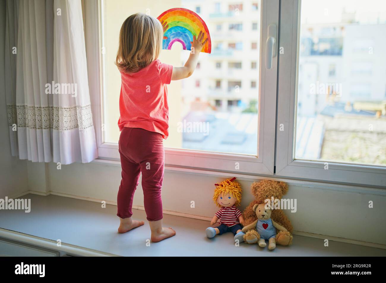 Adorable toddler girl attaching rainbow drawing to window glass as sign ...