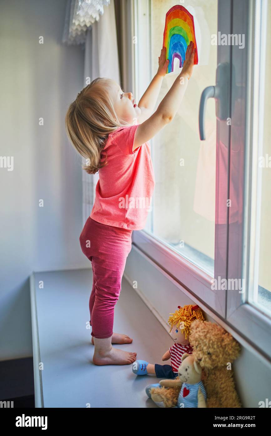 Adorable toddler girl attaching rainbow drawing to window glass as sign ...
