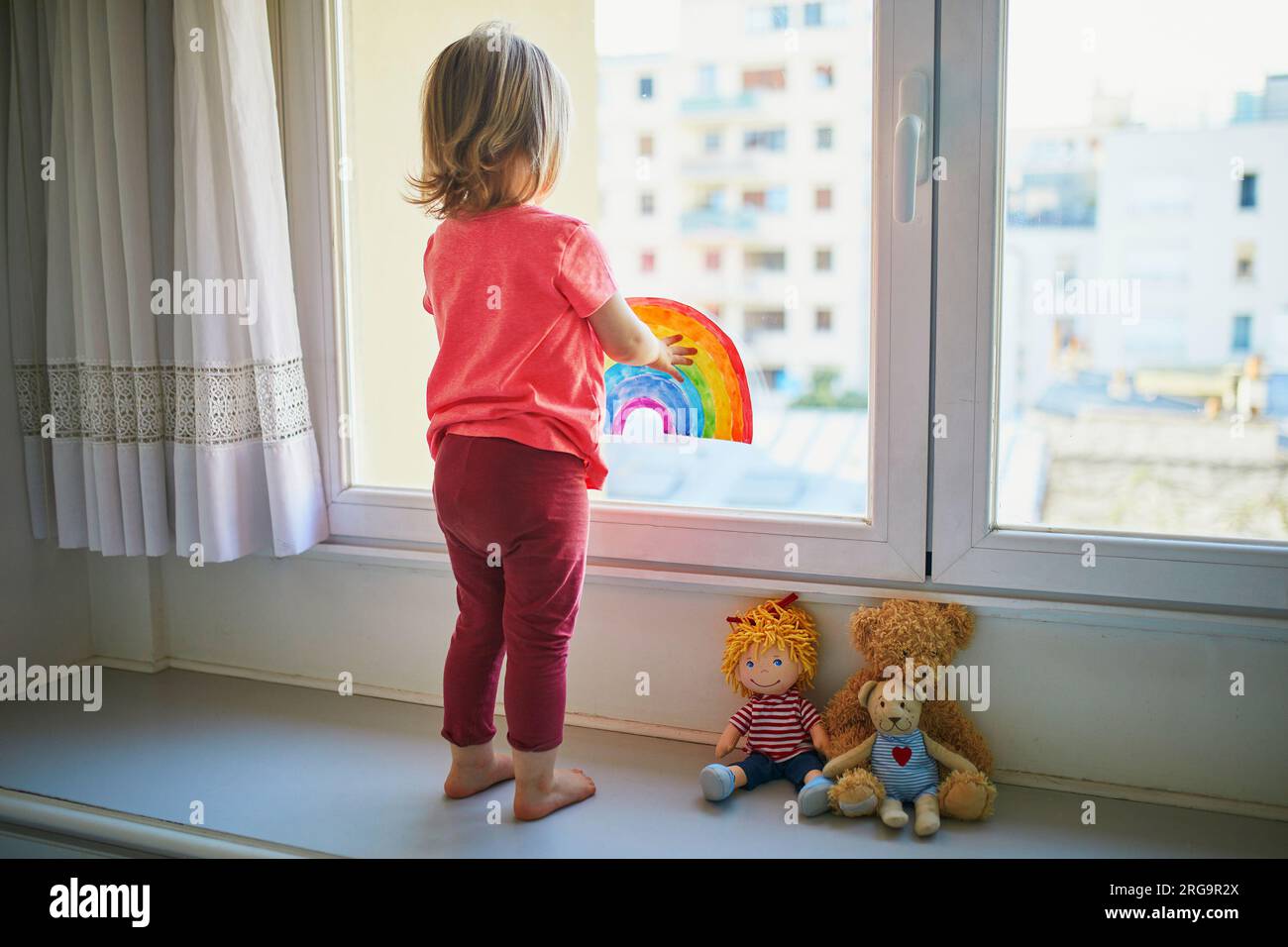 Adorable toddler girl attaching rainbow drawing to window glass as sign of hope. Creative games ...