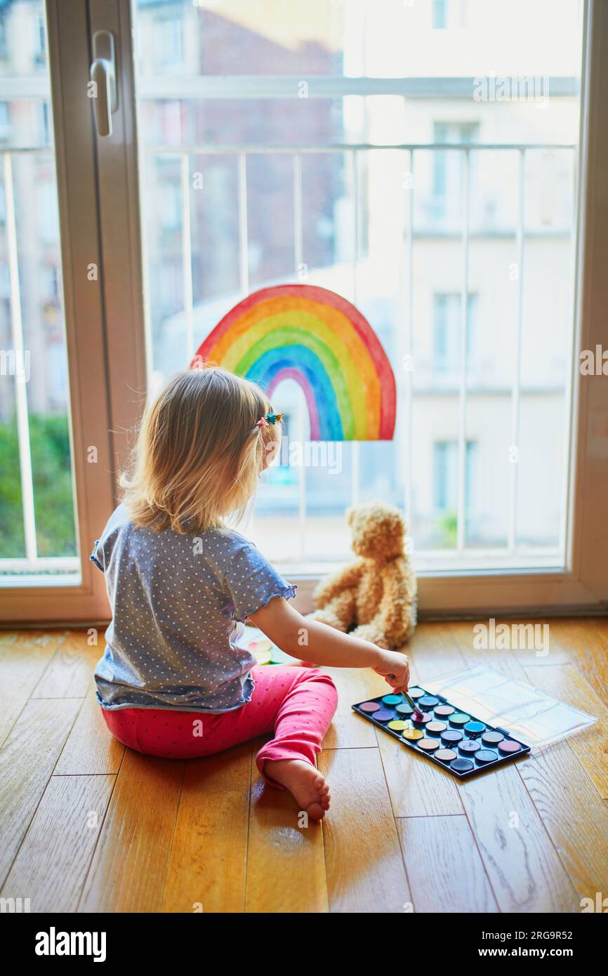 Adorable toddler girl painting rainbow on the window glass as sign of hope. Creative games for ...