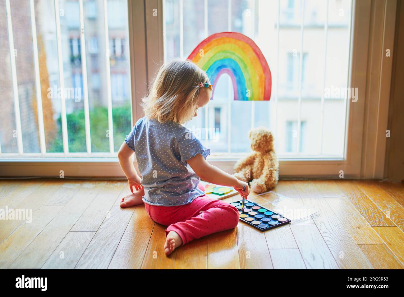 Adorable toddler girl painting rainbow on the window glass as sign of hope. Creative games for ...