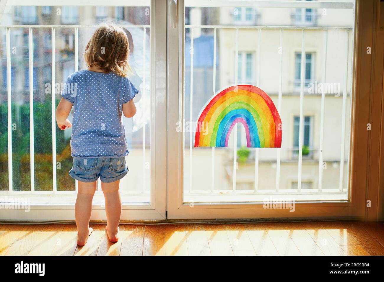 Adorable toddler girl looking through the window with rainbow drawing ...