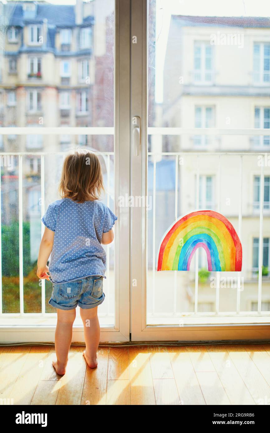 Adorable toddler girl looking through the window with rainbow drawing ...