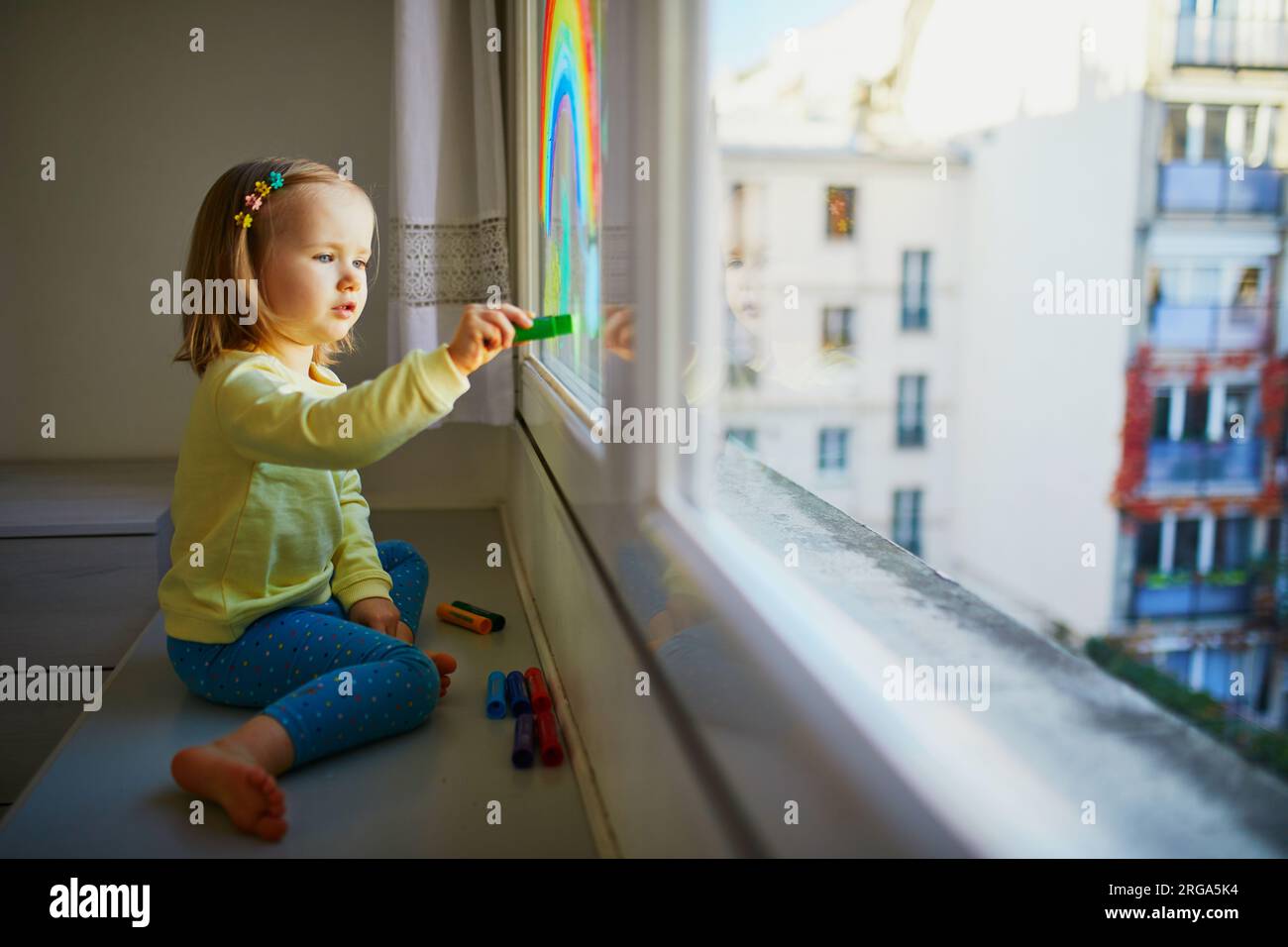 Adorable toddler girl drawing rainbow on window glass as sign of hope ...