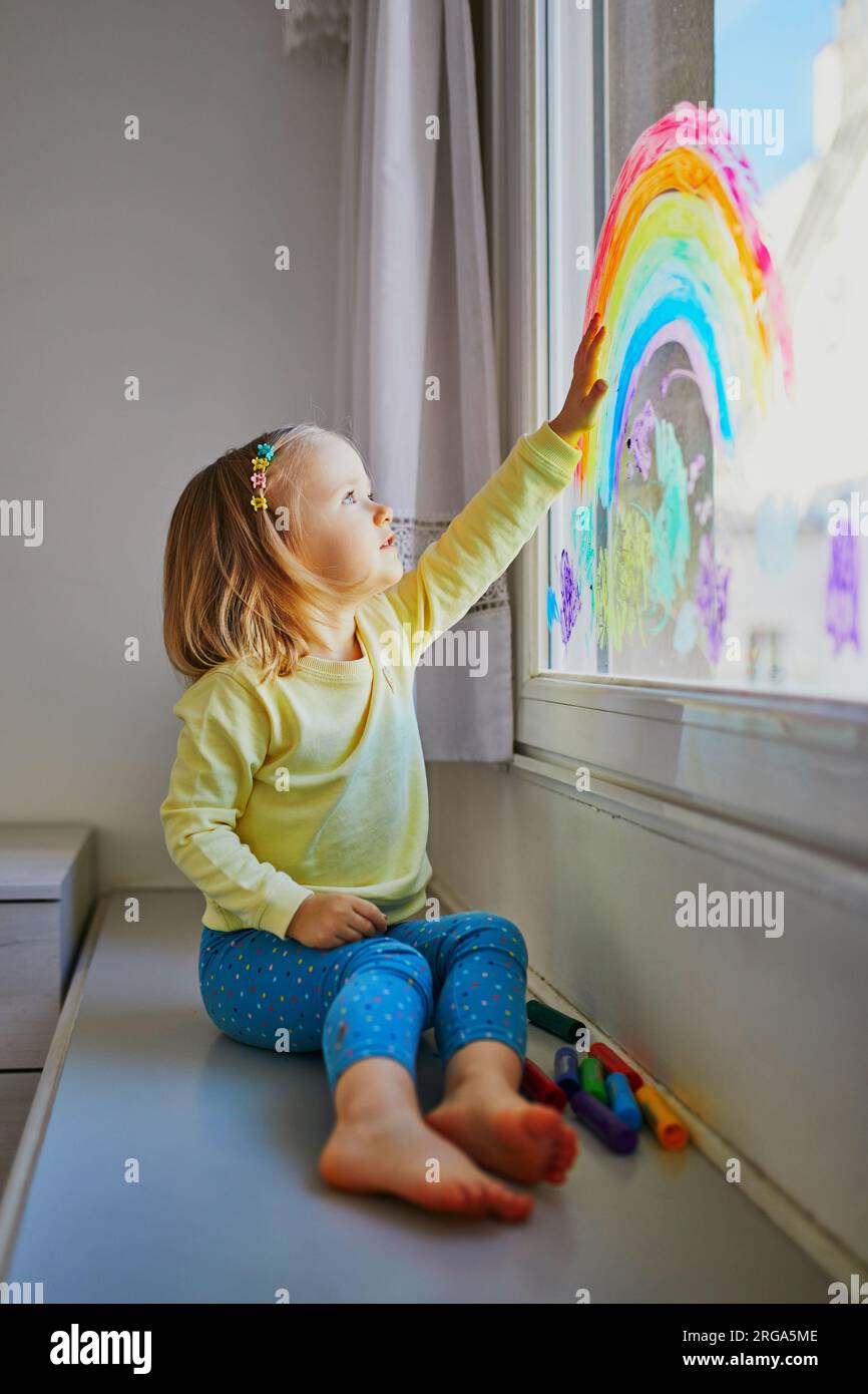 Adorable toddler girl drawing rainbow on window glass as sign of hope ...