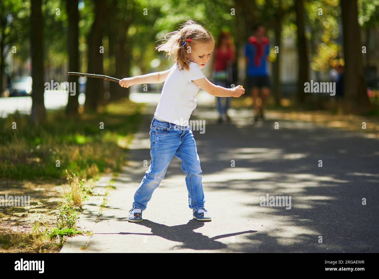 Adorable preschooler girl playing with a stick on a sunny street ...