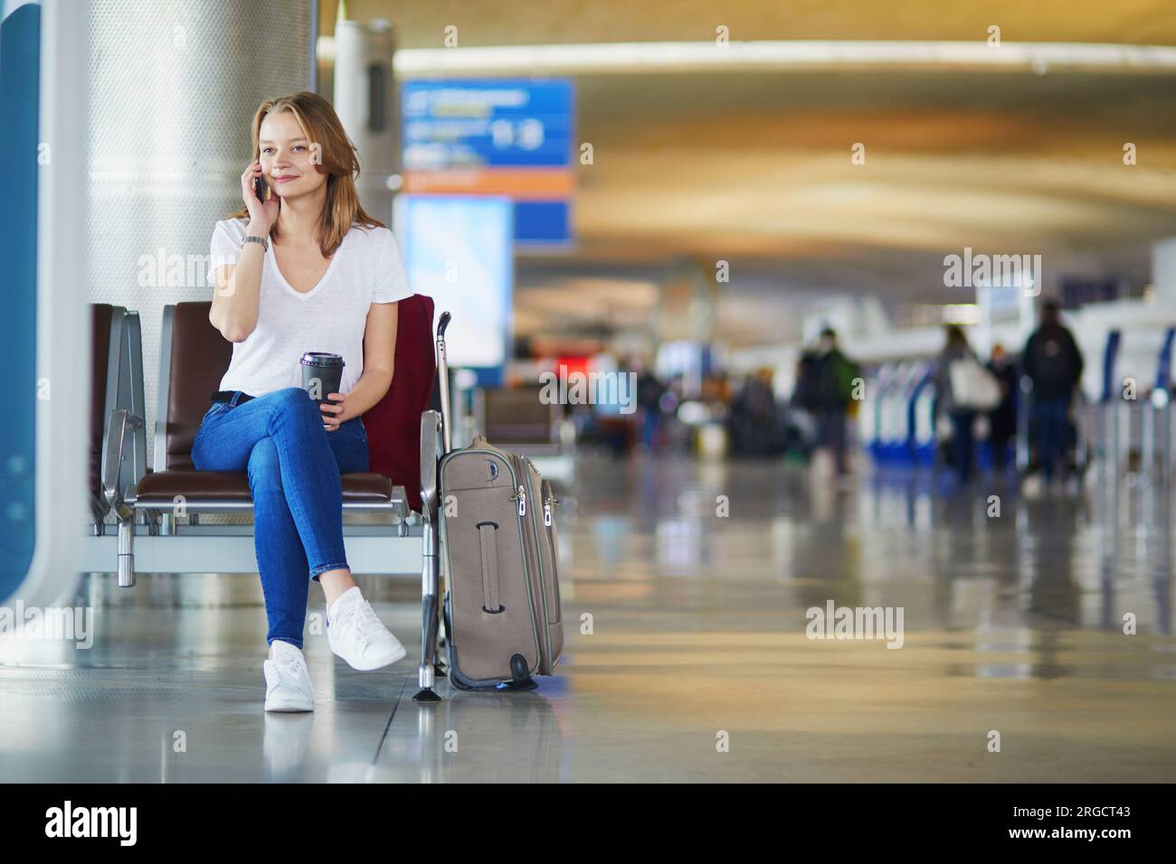 Young woman in international airport with luggage and coffee to go, waiting for her flight and ...
