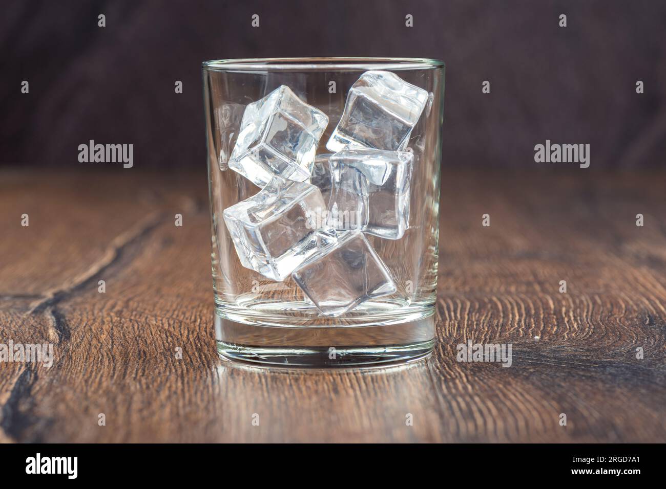 Ice cubes in an empty glass on a wooden bar counter, dark background. Fake or artificial acrylic ...