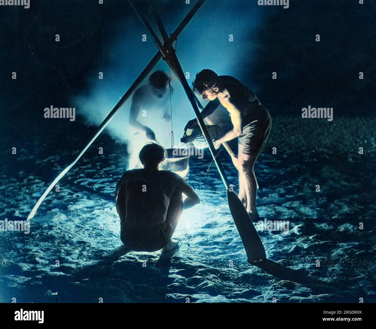 Three young men cooking a meal on a beach from a cauldron hanging from ...