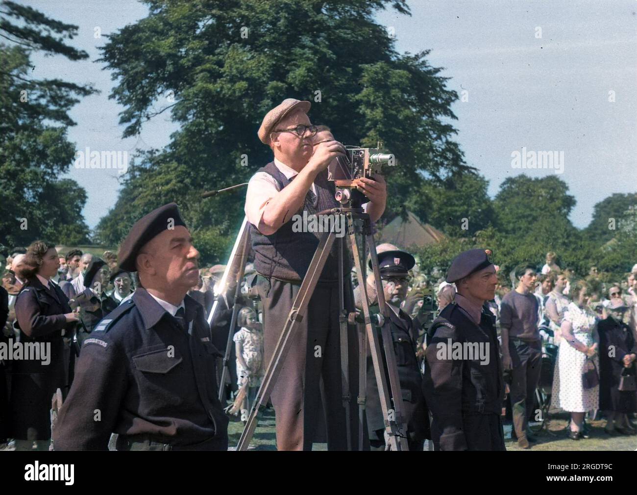 People in a park, watching an event. A man stands with a cine camera on ...
