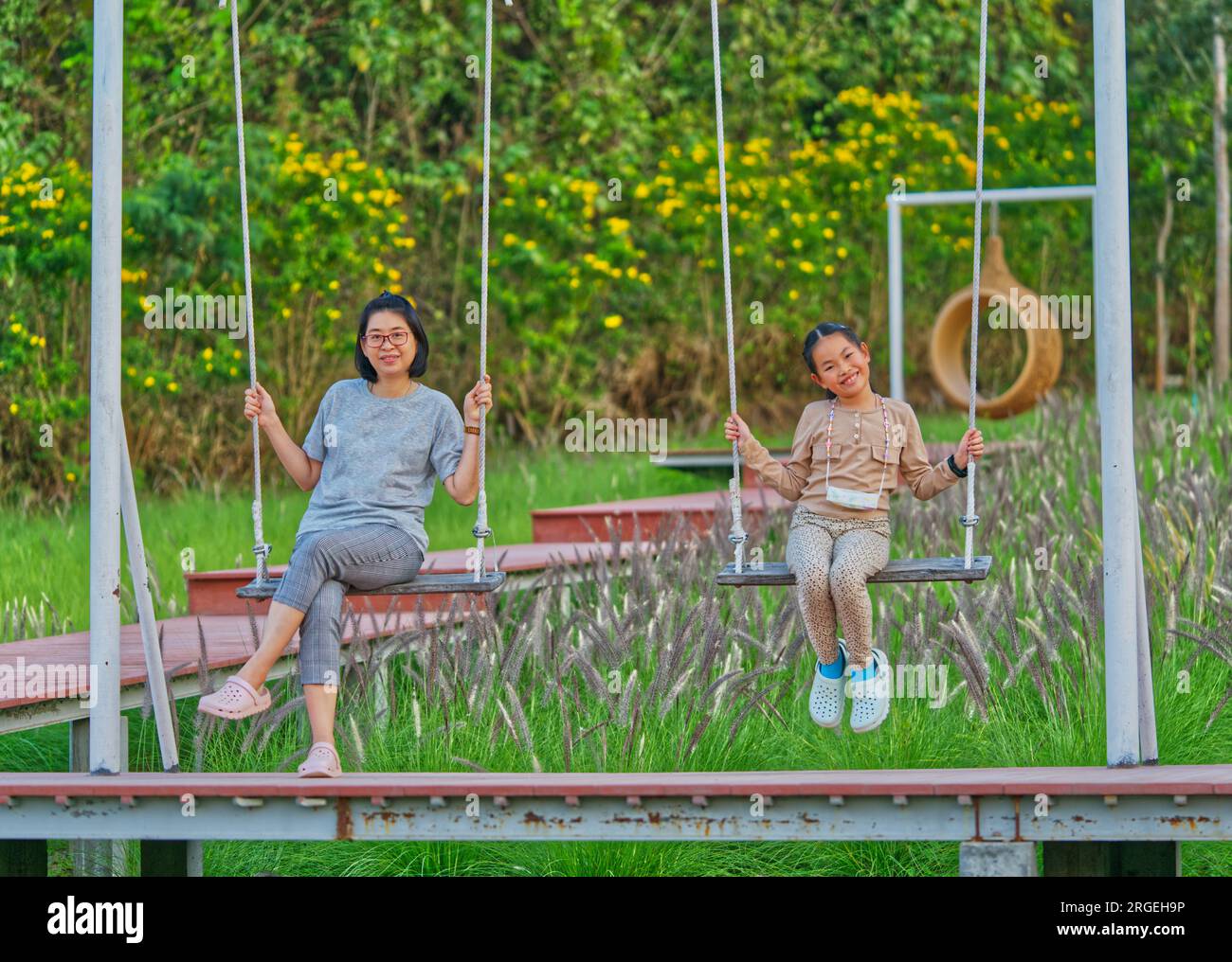 Asian middle-aged mother sitting on a wooden swing in the garden with a little child girl or ...