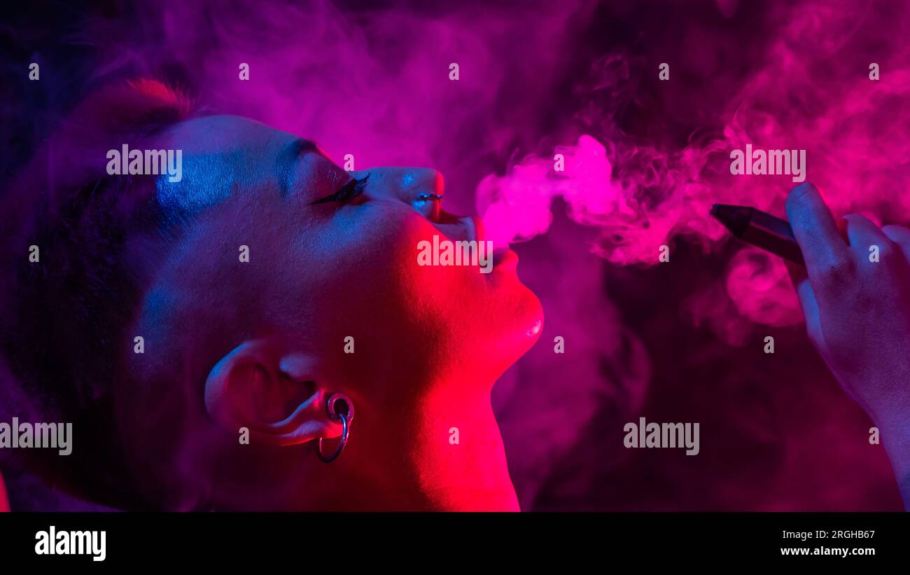 Portrait asian woman with shaved temples smokes vape in neon light ...