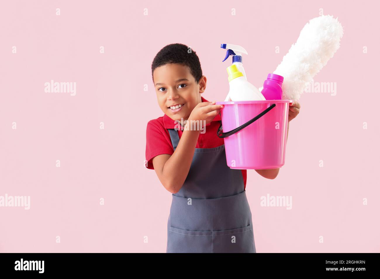 Little African-American boy with cleaning supplies in bucket on pink ...