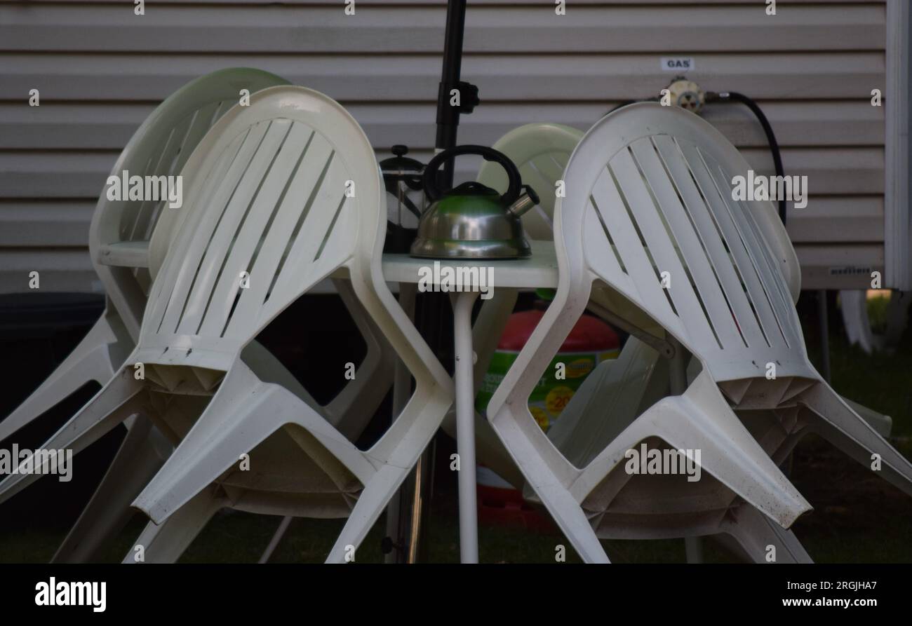 On a rainy campsite, 4 white plastic garden chairs lean on a table ...