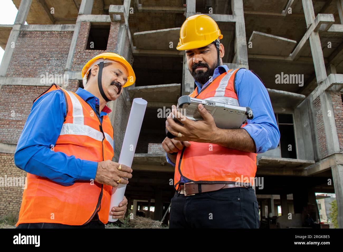 Two Indian male civil engineers or architect wearing helmet and vest holding digital tablet ...