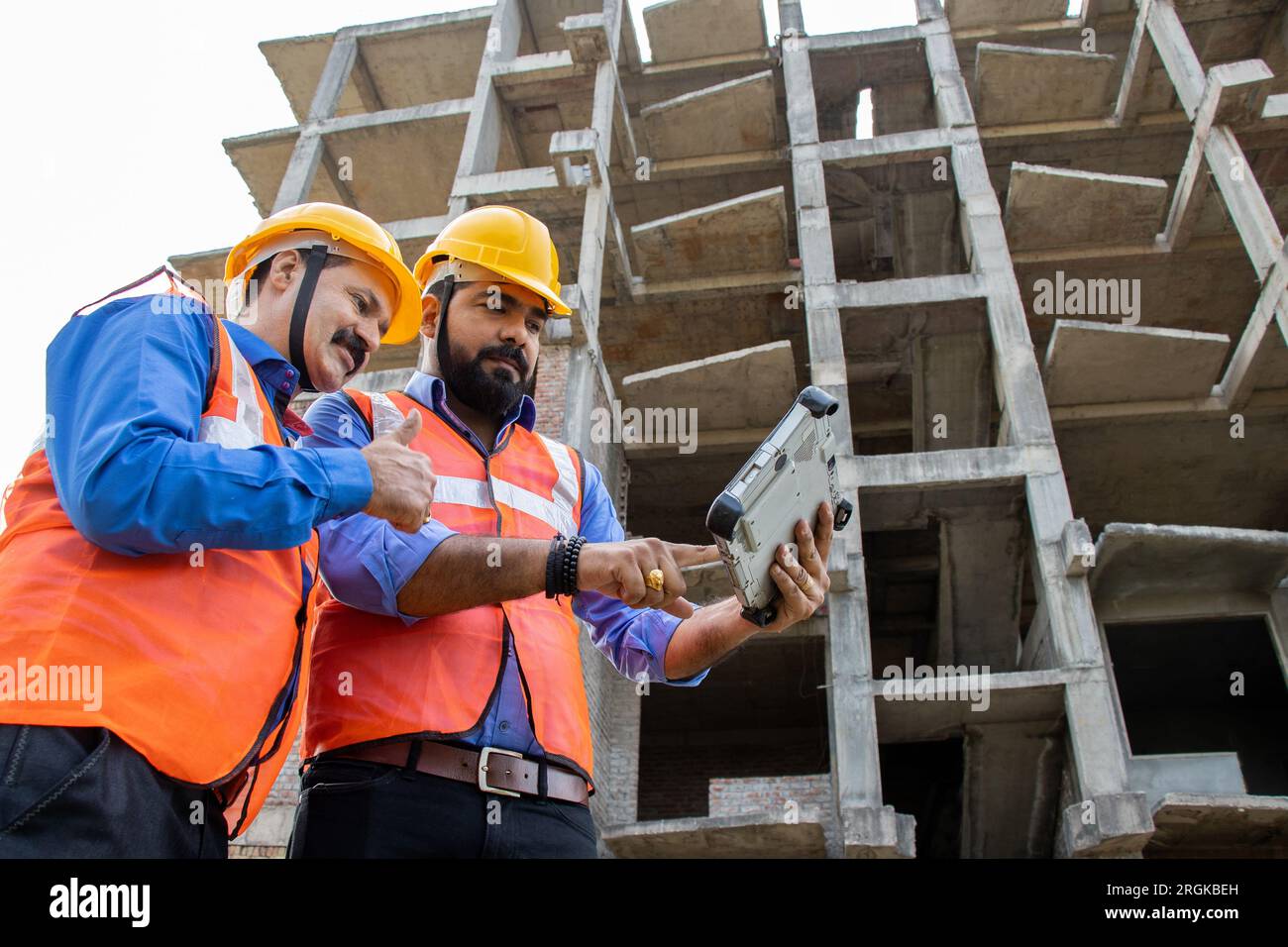 Two Indian male civil engineers or architect wearing helmet and vest holding digital tablet ...