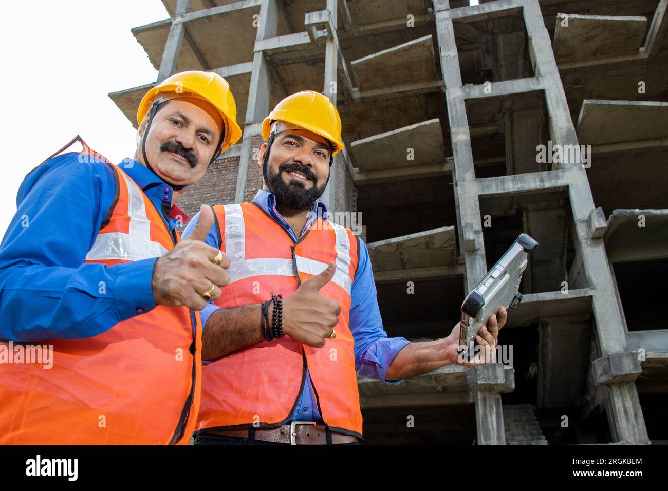 Two Indian male civil engineers or architect wearing helmet and vest holding digital tablet ...