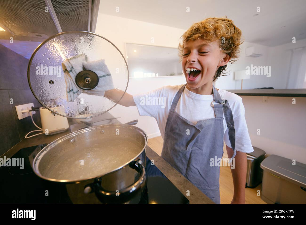 Positive child in apron with lid against boiling water in saucepan on ...