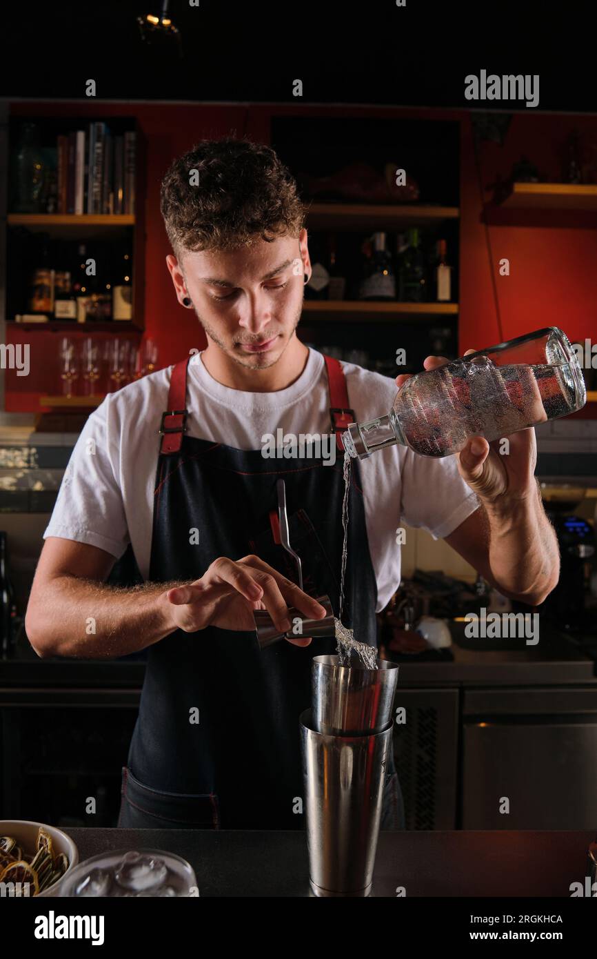 Young male bartender with earrings standing near counter and pouring vodka from bottle and syrup ...
