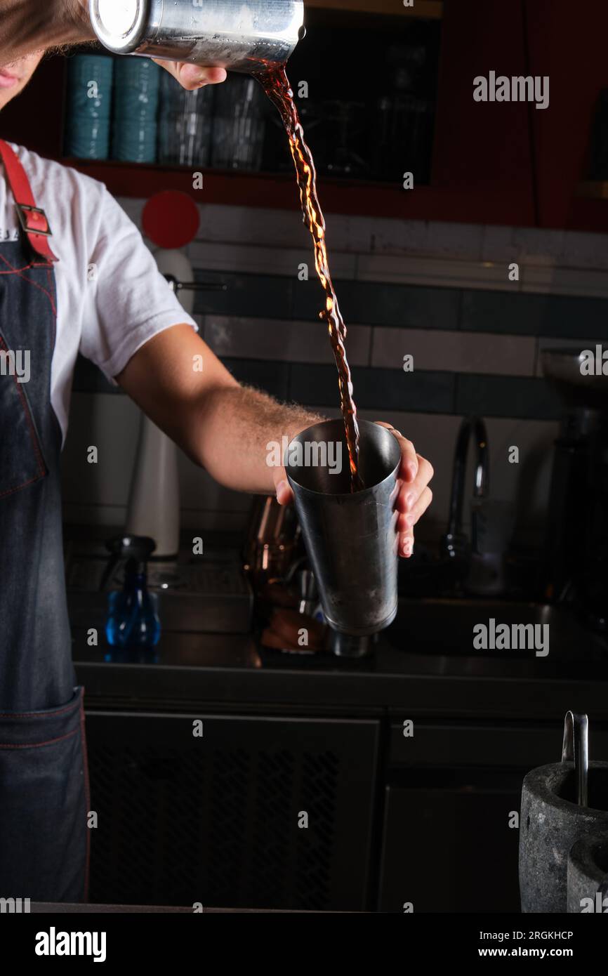Professional male bartender in apron pouring alcohol drink into shaker while mixing cocktail ...