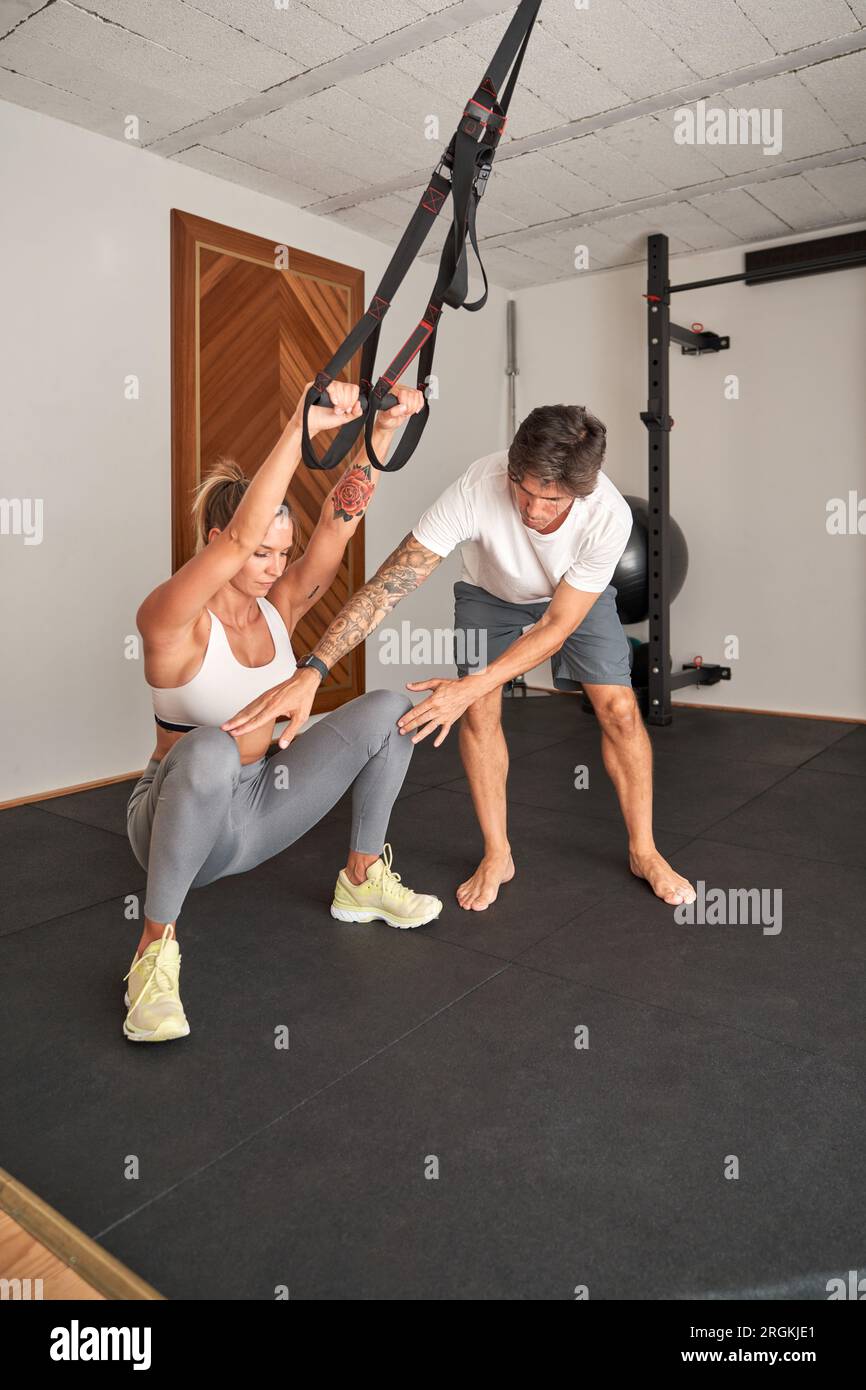 Full body of sporty young woman doing squats with TRX ropes with ...