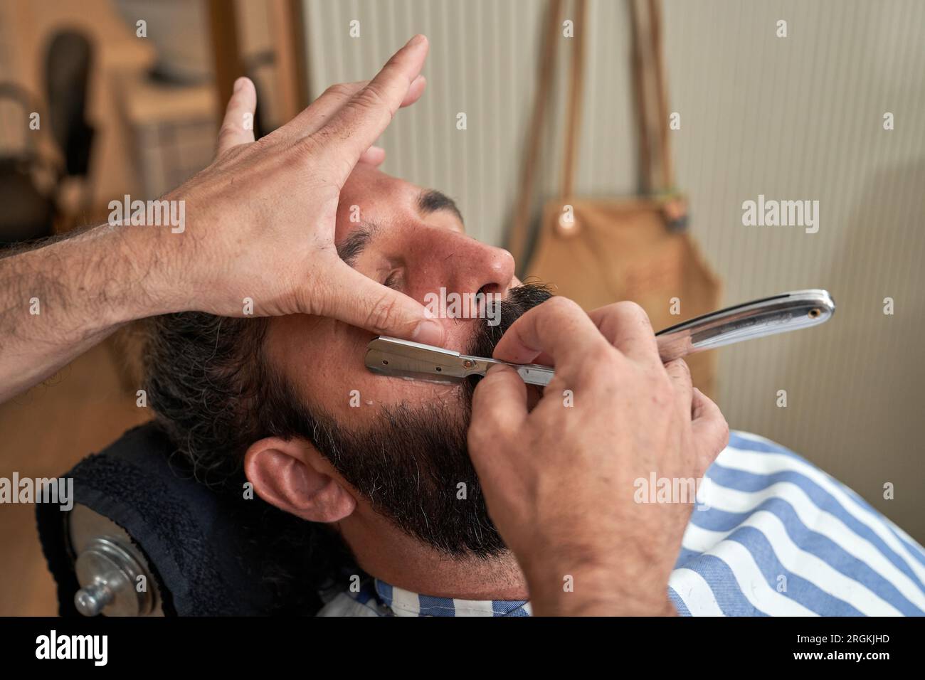Crop barber with tattoo cutting beard of client with straight blade ...