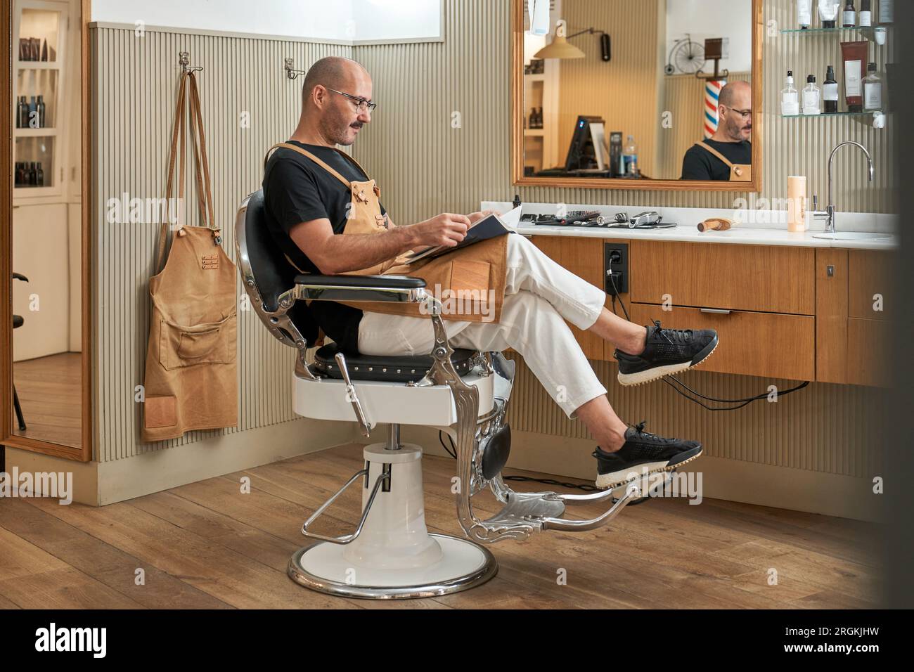 Side view of mature male barber in apron sitting on chair and reading ...