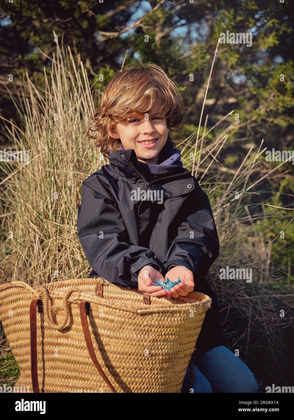 Smiling kid with curly blond hair sitting on ground near wicker bag and ...