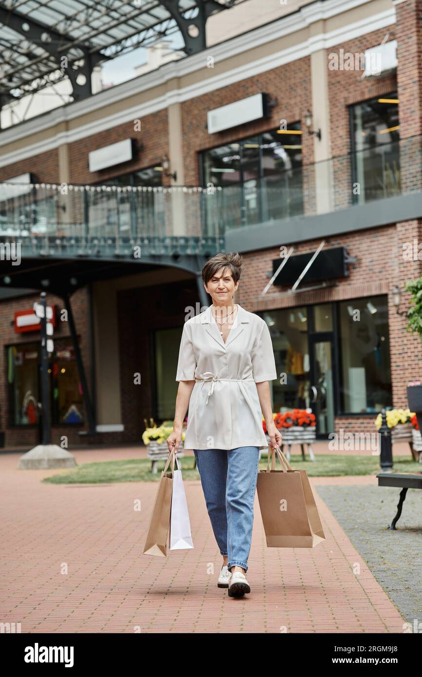 pleased senior woman walking with shopping bags on urban street, retail ...