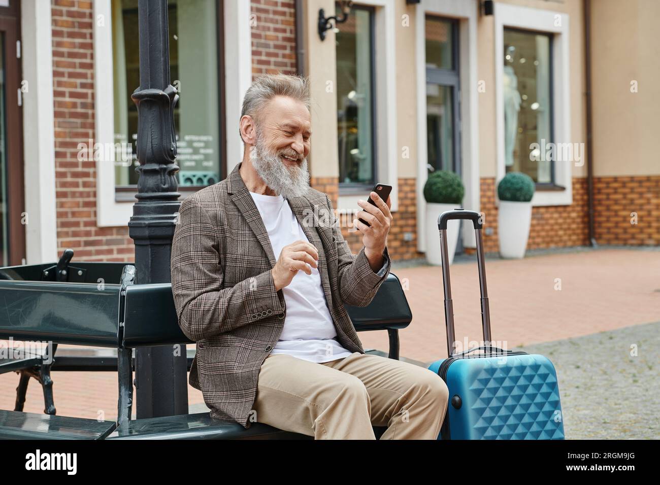 happy senior man using smartphone and sitting on bench near luggage ...