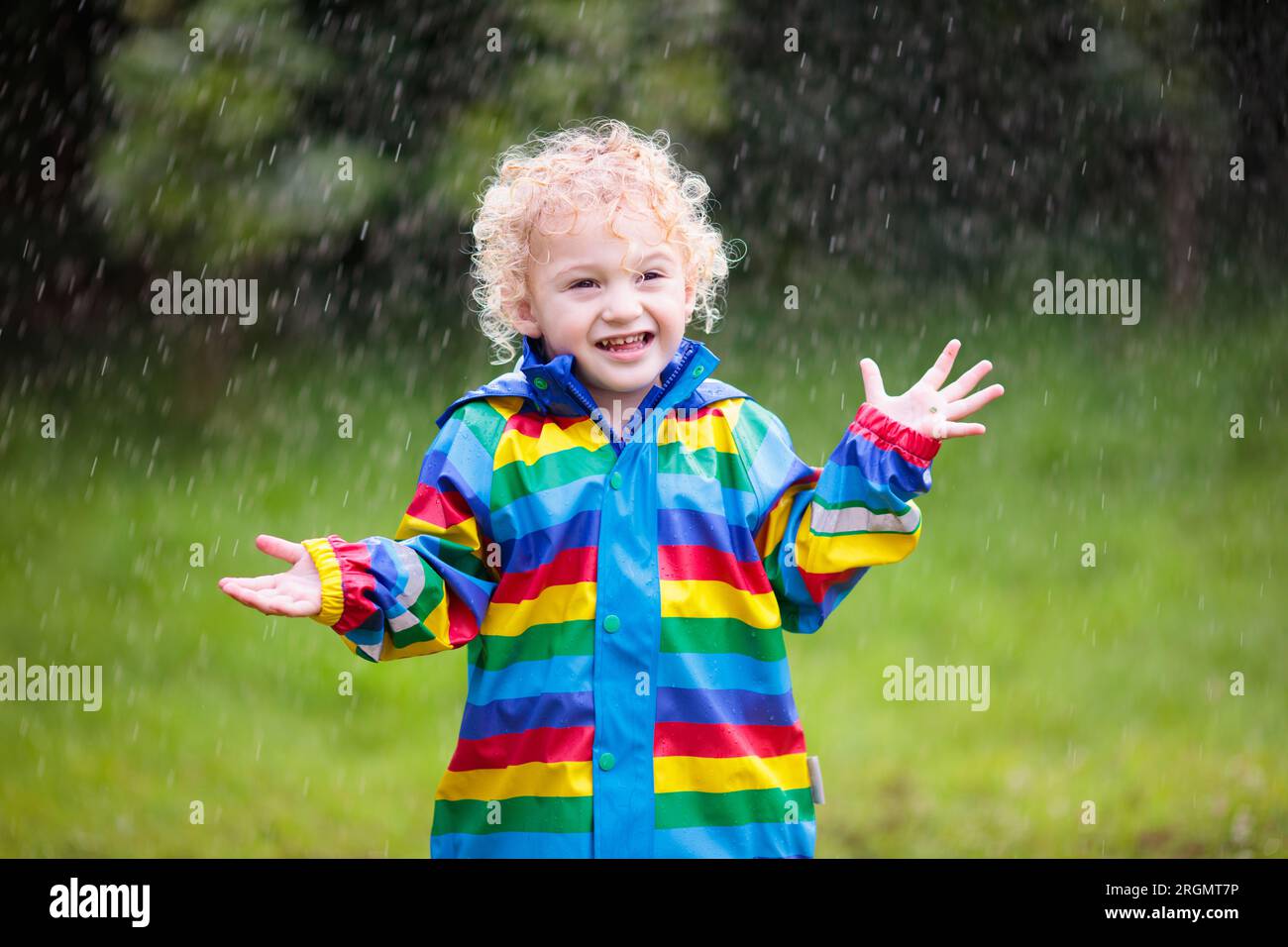 Little blond curly boy in a waterproof jacket in rainbow color playing ...
