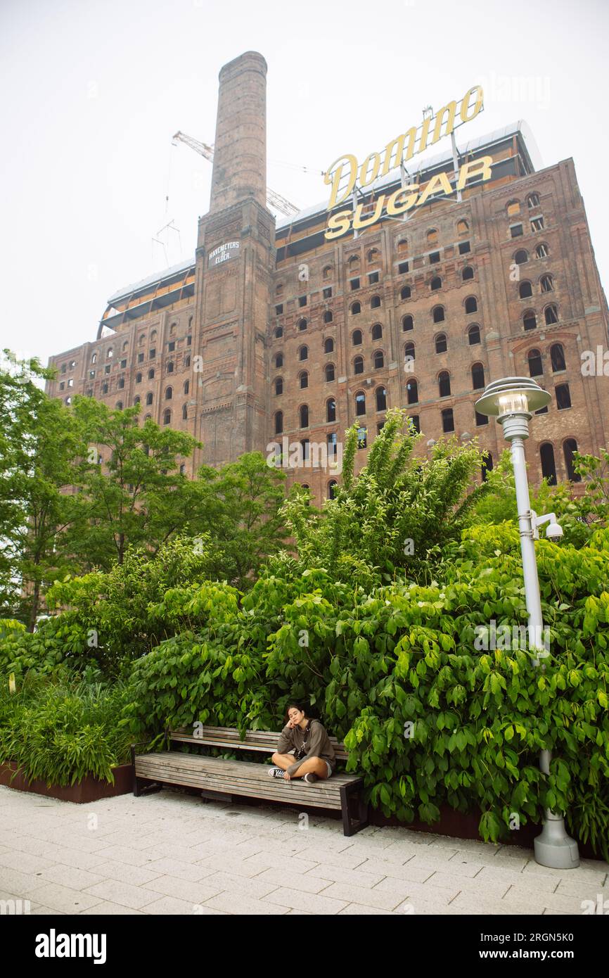 Teenager girl sitting on a bench with Domino Sugar building at the back ...