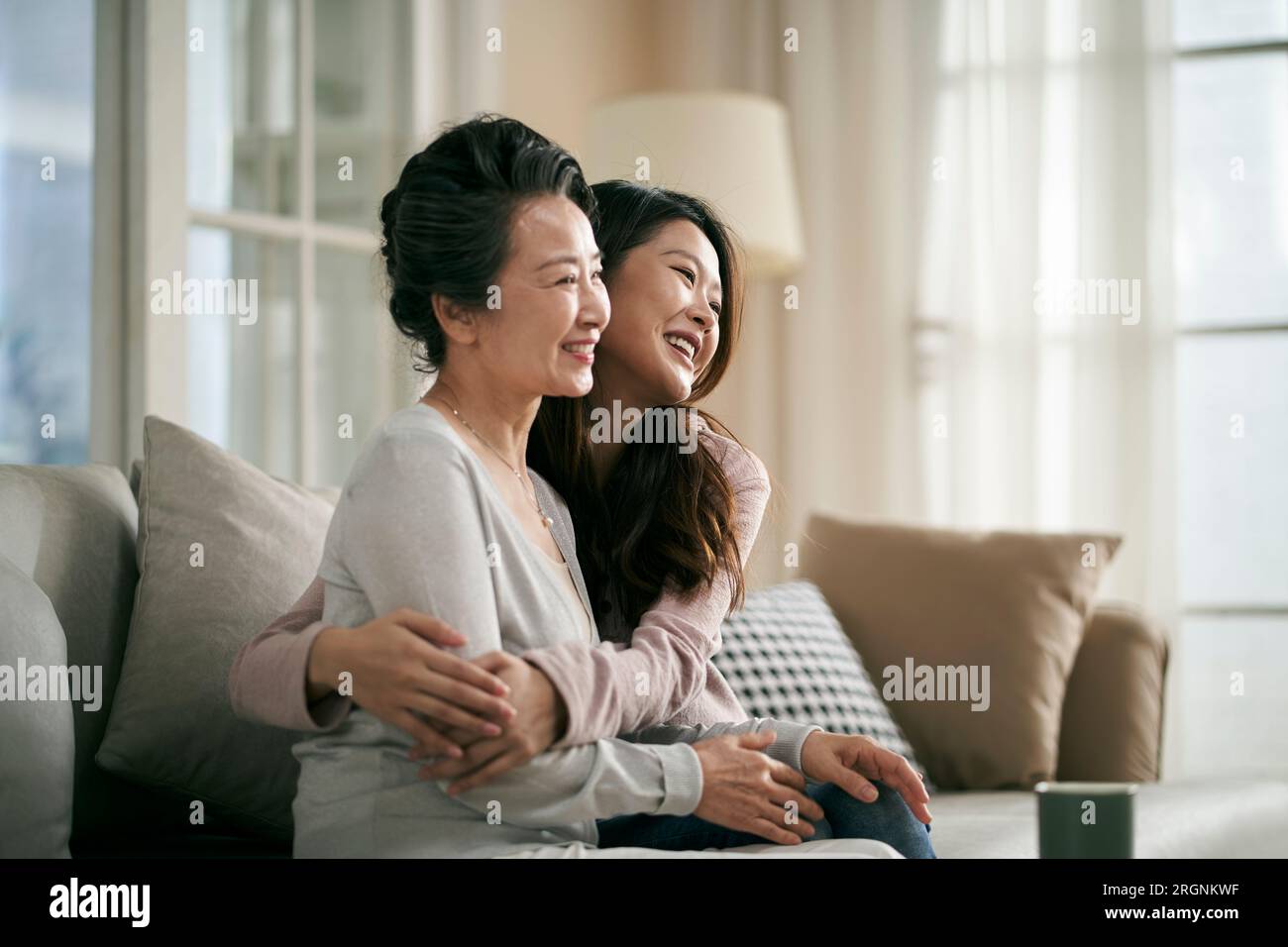 loving asian adult daughter sitting on couch at home hugging senior mother happy and smiling ...