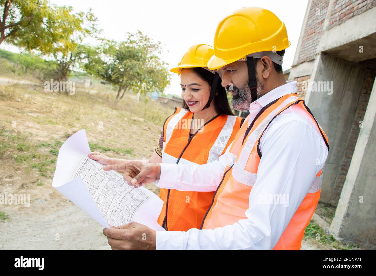 Two Indian male and female civil engineers or architect wearing helmet and vest holding ...