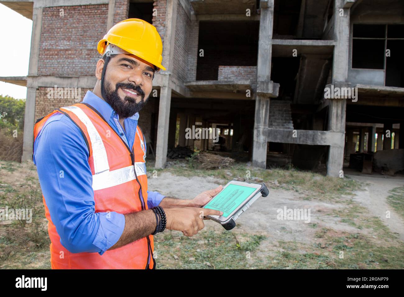Young Indian male civil engineer or architect wearing helmet and vest holding digital tablet ...