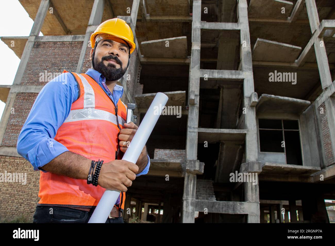 Young Indian male civil engineer or architect wearing helmet and vest holding paperwork ...