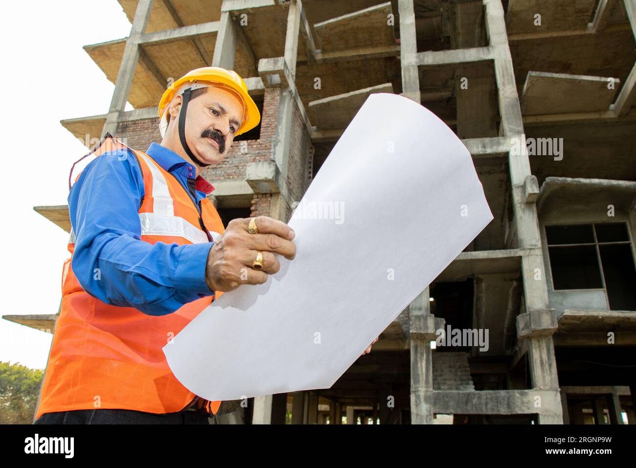 Mature Indian male civil engineer or architect wearing helmet and vest holding paperwork ...