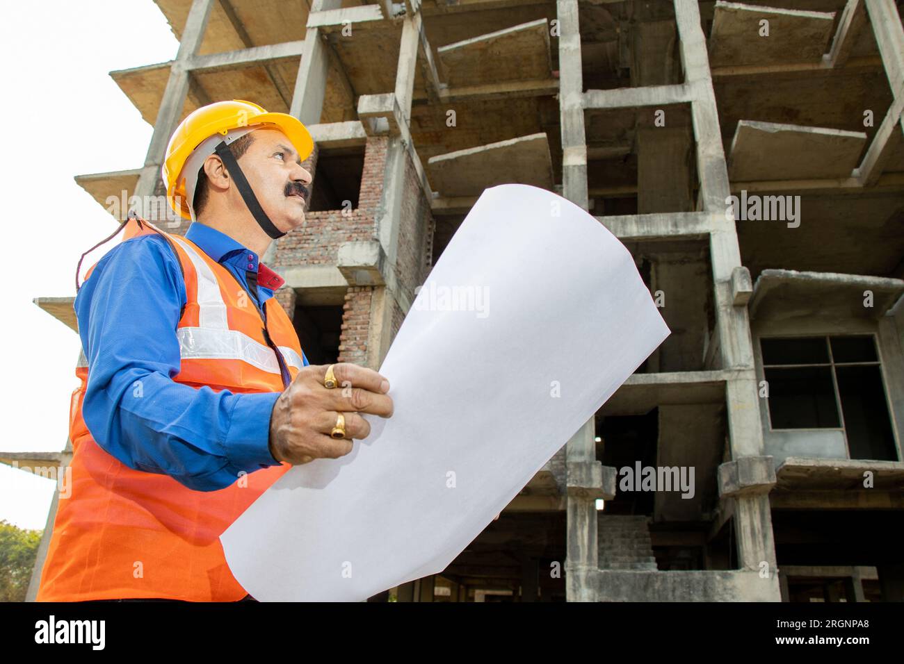 Mature Indian male civil engineer or architect wearing helmet and vest holding paperwork ...