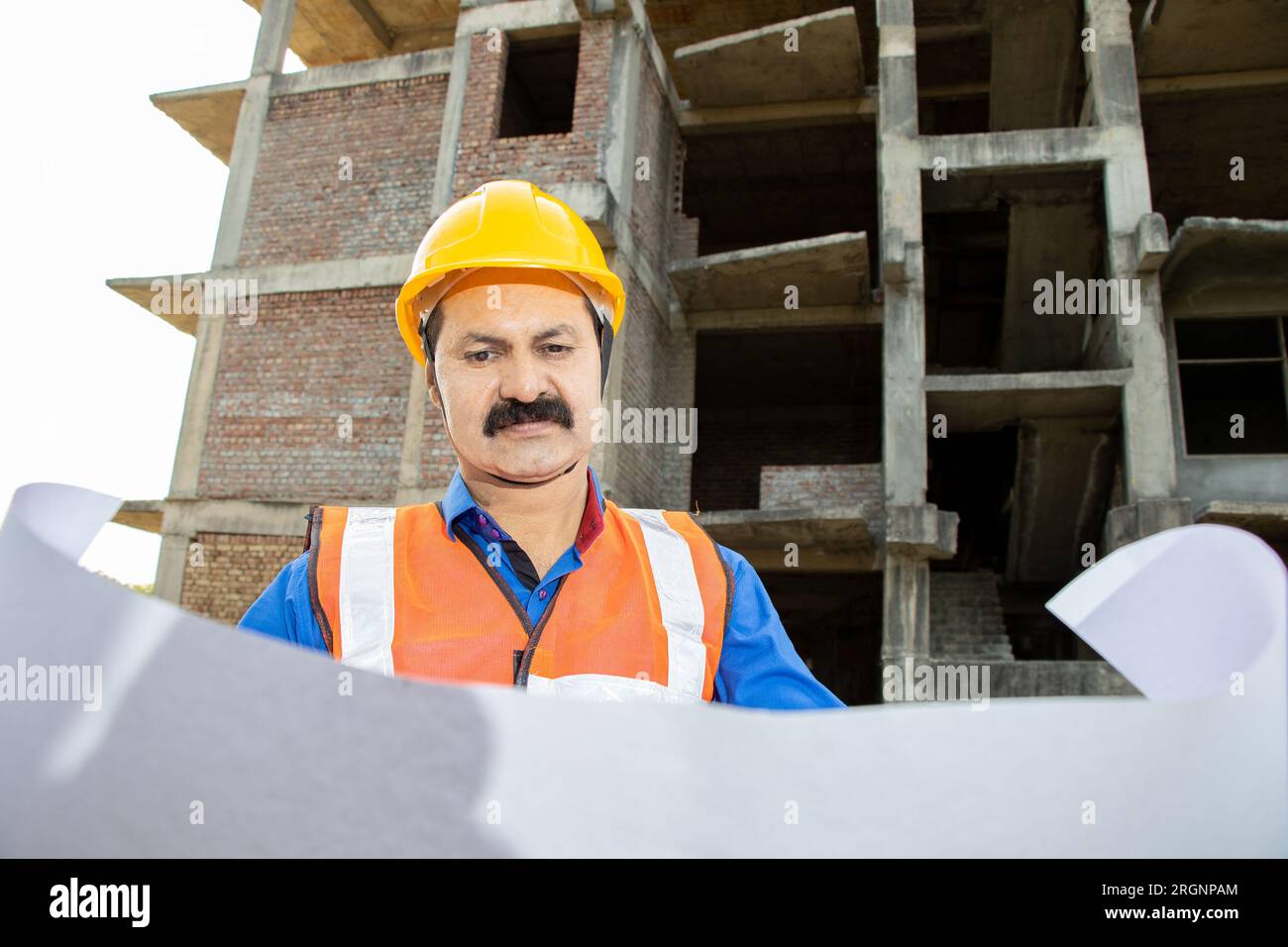 Mature Indian male civil engineer or architect wearing helmet and vest holding paperwork ...