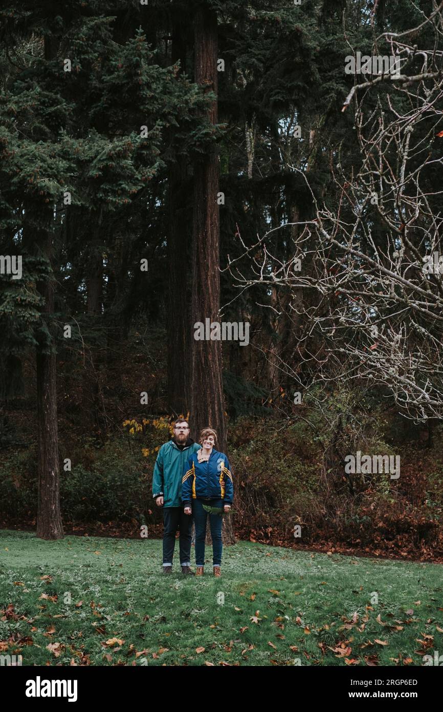 Young couple in retro windbreakers pose deadpan in front of pine trees ...