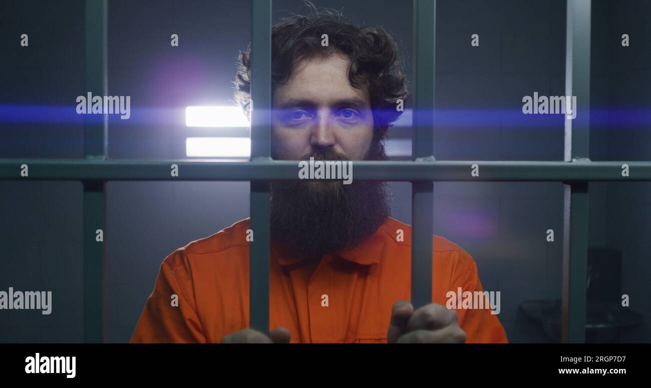 Prisoner in orange uniform holds metal bars, stands in prison cell in ...