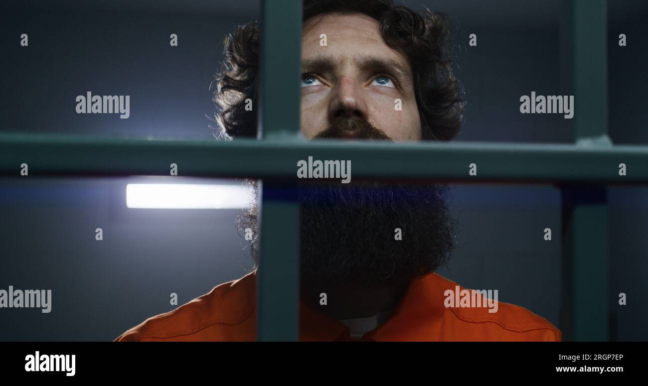 Male criminal in orange uniform holds metal bars with handcuffed hands ...