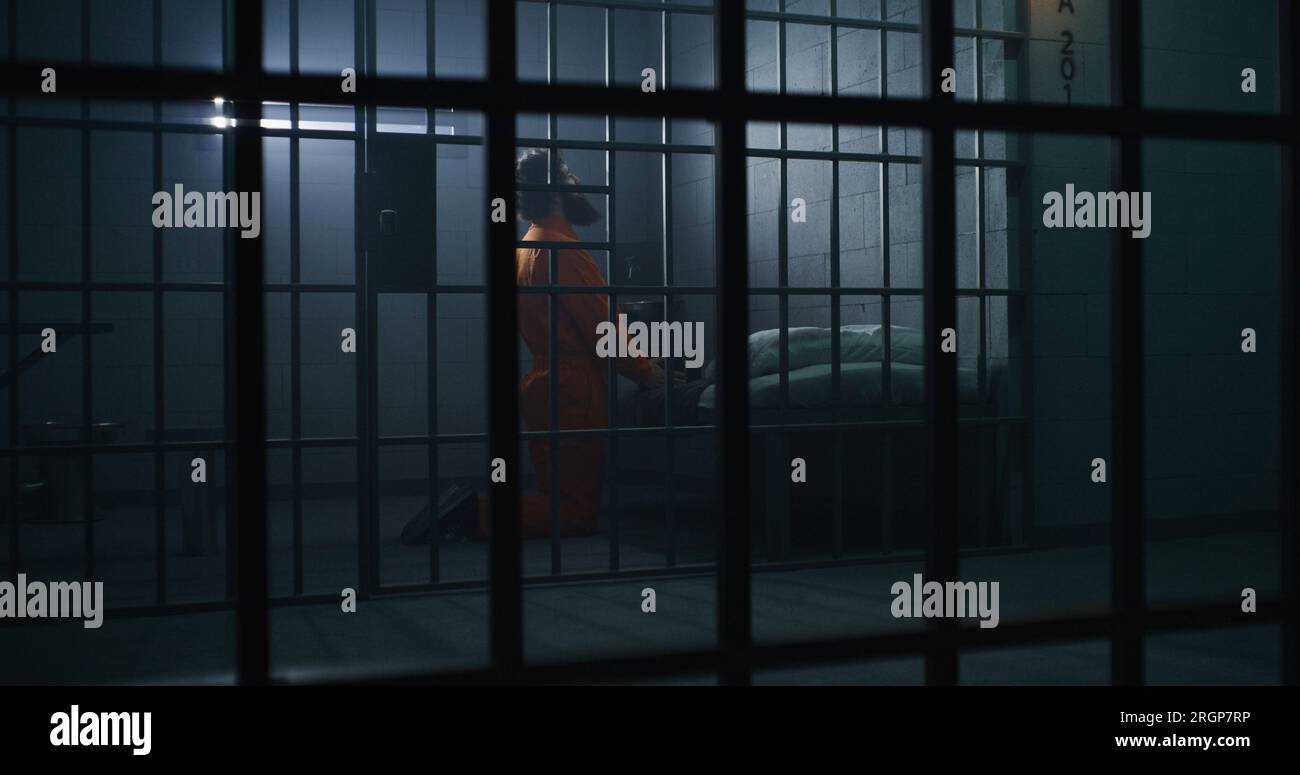 Prisoner in orange uniform kneels near bed, prays to God in prison cell ...
