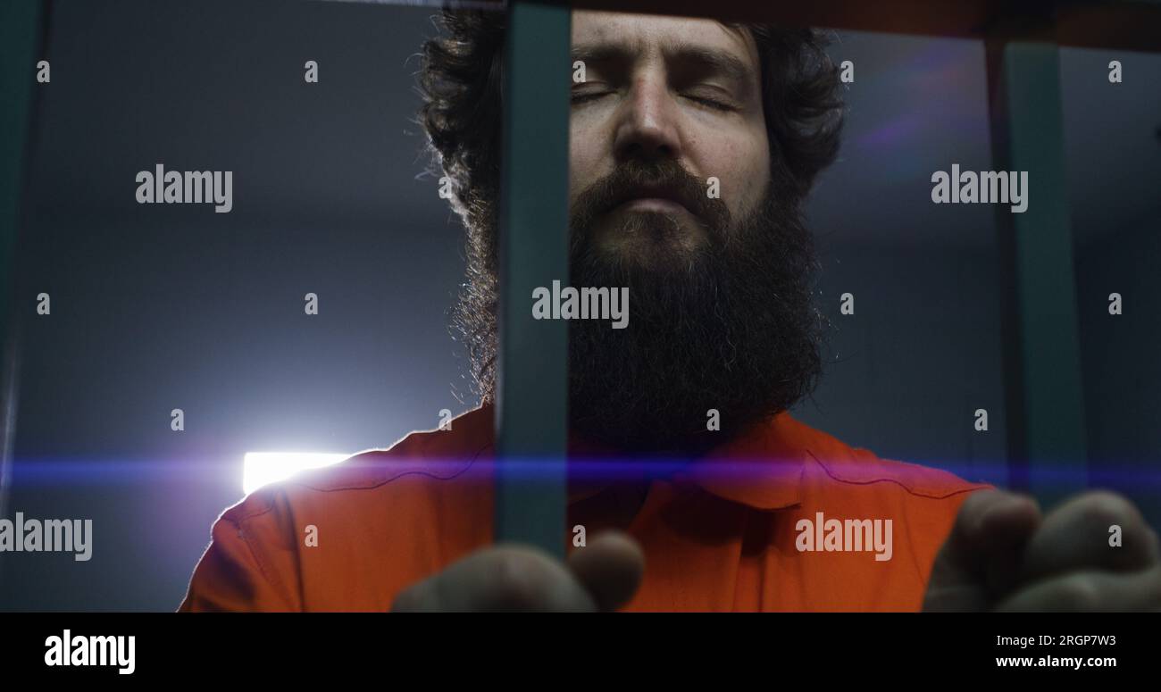 Male criminal in orange uniform holds metal bars with handcuffed hands ...