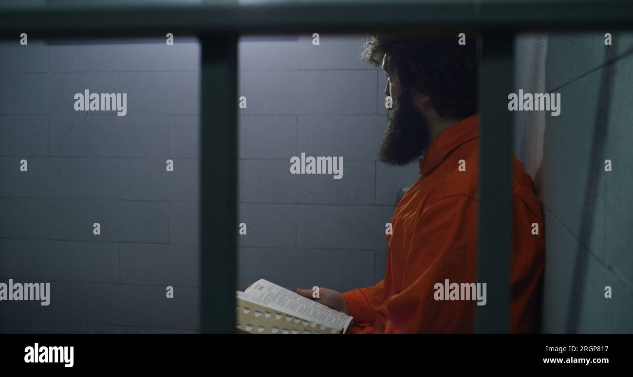 Male prisoner in orange uniform sits on the bed, reads Bible, looks at ...