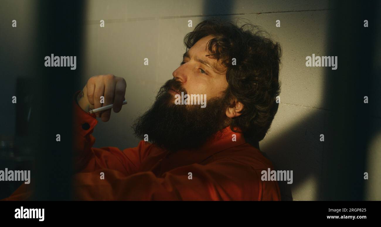 Thoughtful male prisoner in orange uniform sits on the bed, lights ...