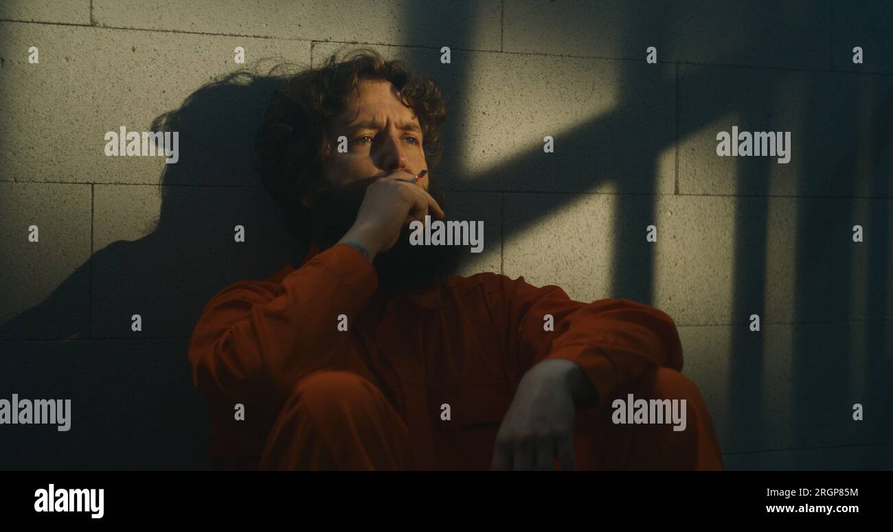 Depressed prisoner in orange uniform sits on bed, smokes cigarette in ...
