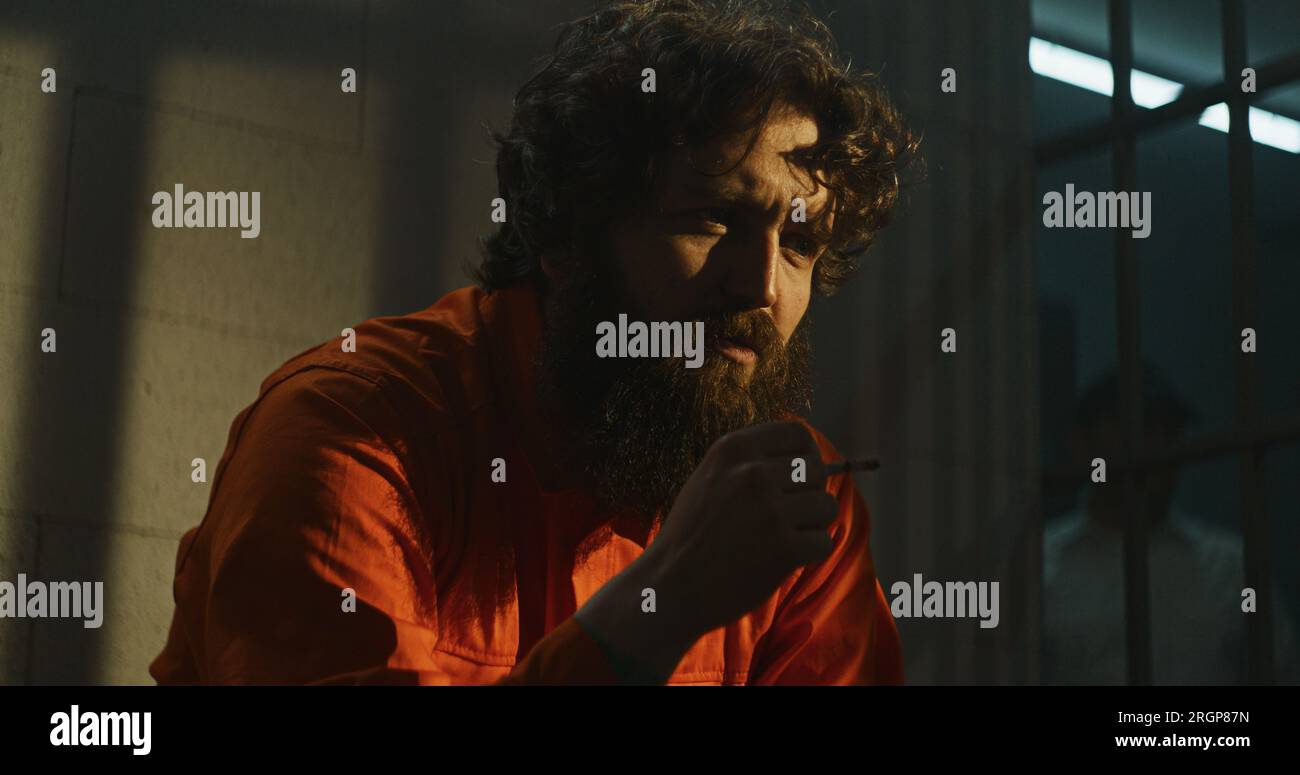 Upset prisoner in orange uniform sits on bed in prison cell, smokes ...