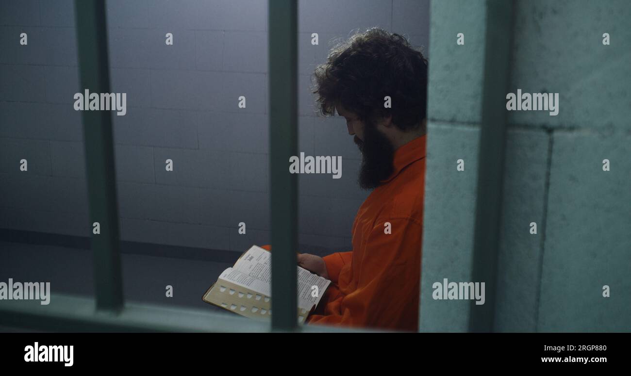 Male prisoner in orange uniform sits on the bed, reads Bible, looks at ...